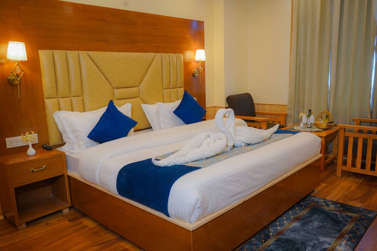 Uma cama de casal com cabeceira, duas mesas de cabeceira, uma cadeira, uma pequena mesa com uma garrafa e uma cortina.