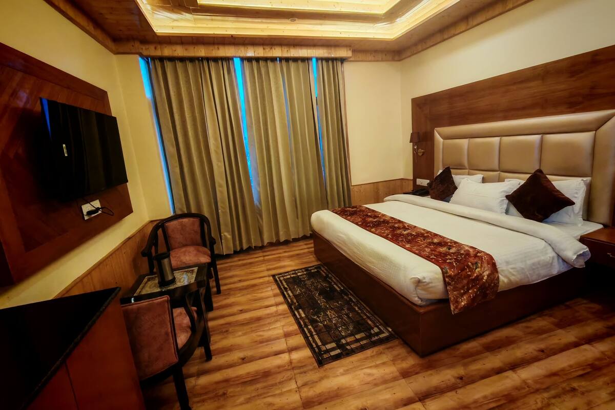Quarto de hotel com piso de madeira, uma cama com cabeceira, televisão fixada na parede, uma escrivaninha com cadeira e uma janela com cortinas.