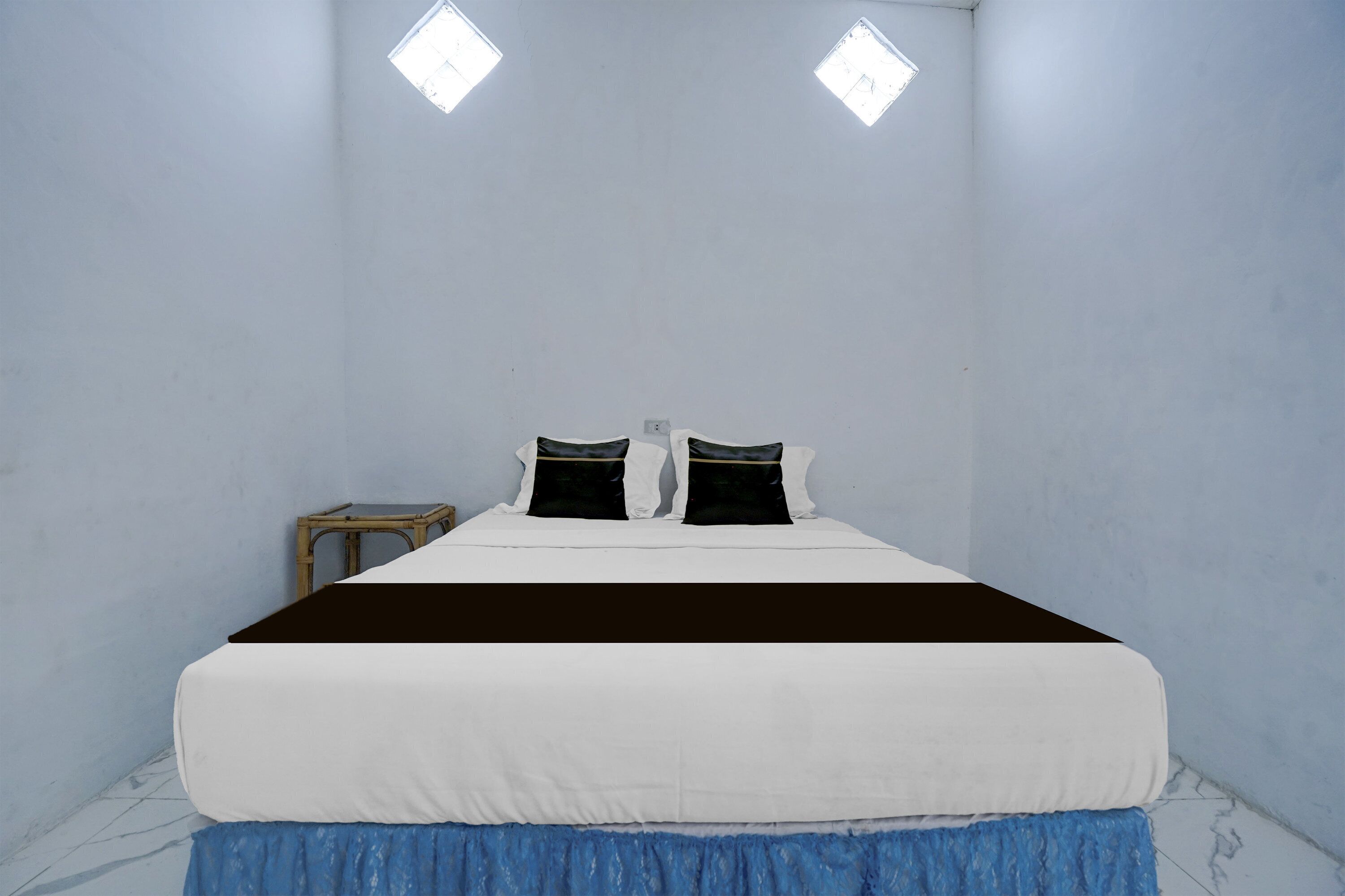 Uma cama com colcha branca e azul, duas almofadas pretas e uma mesinha de cabeceira de madeira.