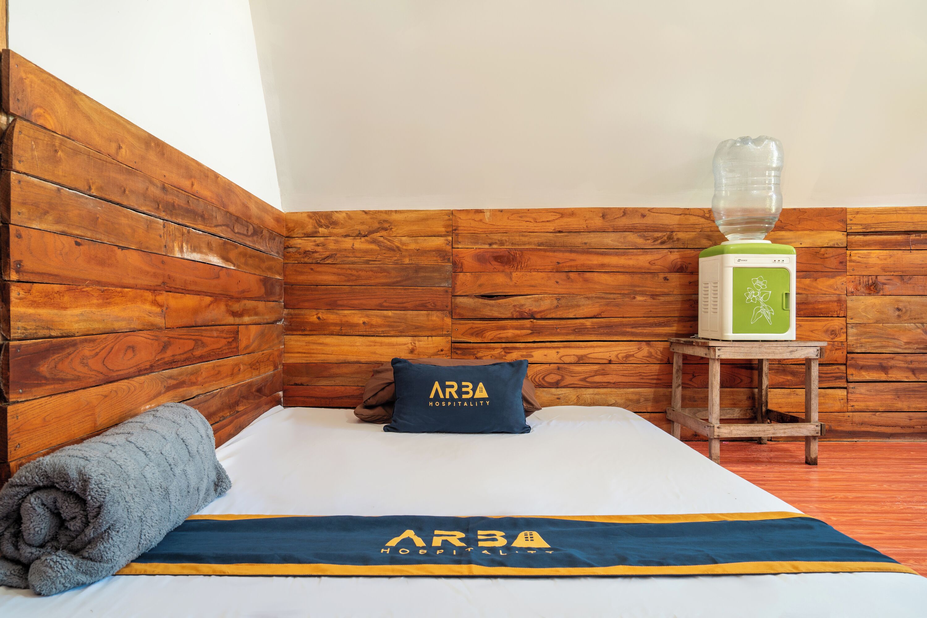 Quarto de hotel com painéis de madeira, uma cama com manta da marca, um travesseiro e um dispensador de água sobre uma mesa de madeira.