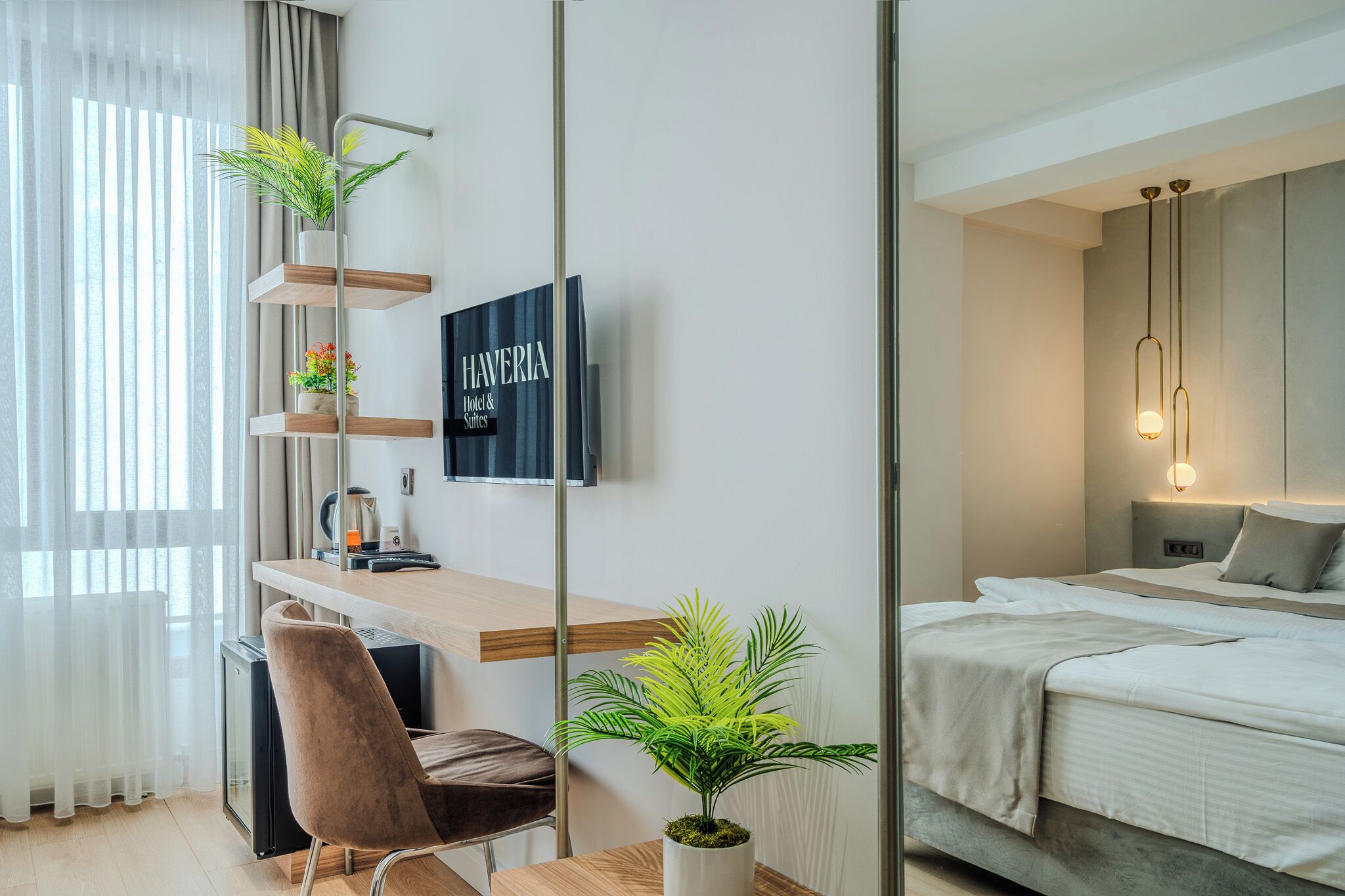 Quarto de hotel moderno com uma cama, uma escrivaninha com cadeira, uma televisão e duas plantas em vasos.