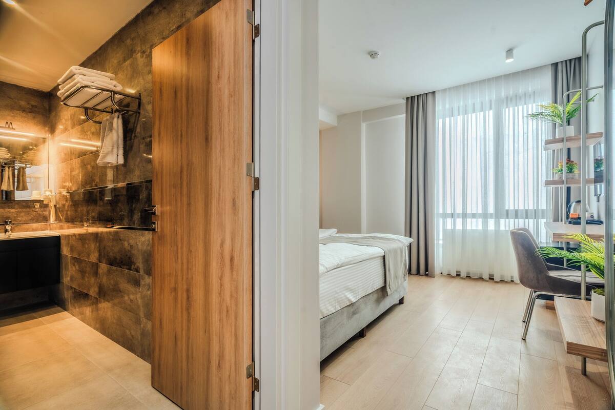 Quarto moderno com cama, escrivaninha e cadeira. Possui banheiro com pia e porta-toalhas.