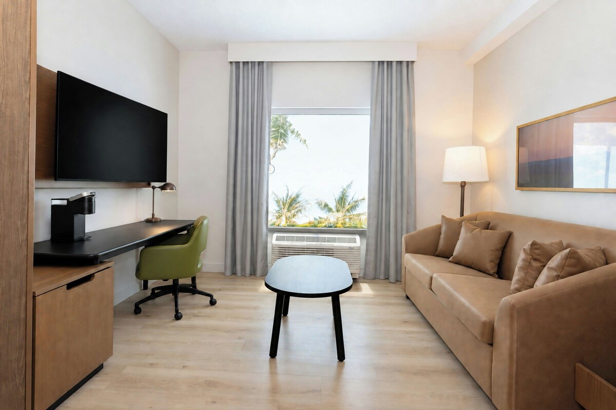Quarto de hotel moderno com televisão de tela plana, escrivaninha com cadeira, sofá, mesinha e janela com cortinas.