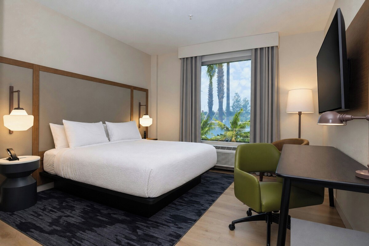 Quarto de hotel com uma cama grande, uma escrivaninha, uma cadeira, televisão e janela com vista para palmeiras.