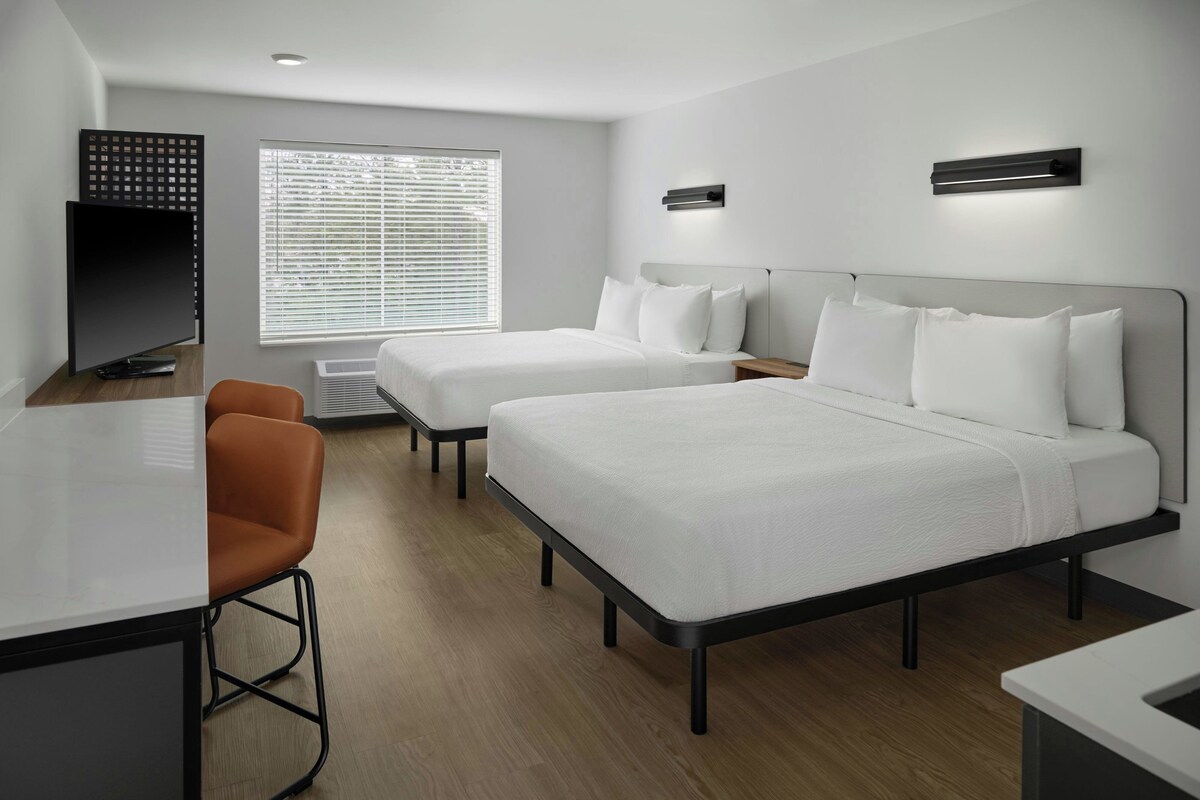 Quarto de hotel com duas camas, uma escrivaninha, uma cadeira e janela com persianas.
