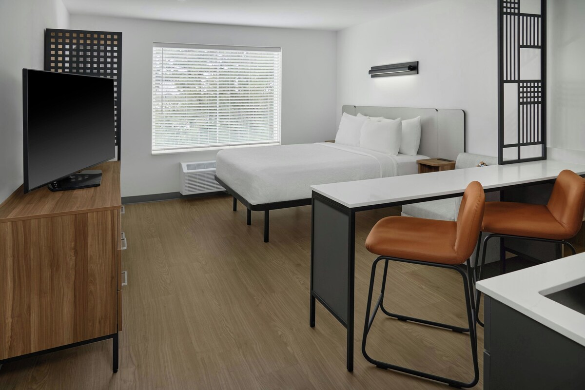 Quarto de hotel moderno com cama, escrivaninha, duas cadeiras, televisão e janela com persianas.