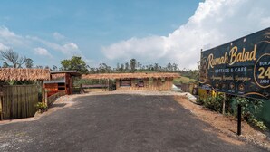 Paisagem rural com um edifício de telhado de colmo e um grande letreiro para 'Pimah Balad COTTAGE & CAFE', com uma estrada de terra que leva a ele.