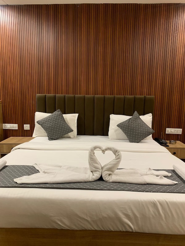 Quarto de hotel com parede revestida em madeira, cama com roupa de cama branca, dois travesseiros, arranjo de toalha em formato de coração e mesa de cabeceira com telefone e abajur.