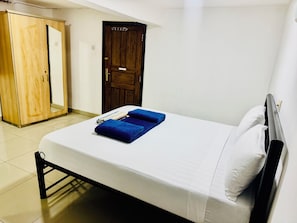 Uma cama bem arrumada com almofadas e um cobertor dobrado, um guarda-roupa de madeira e uma porta em um quarto com paredes brancas e chão de azulejos.