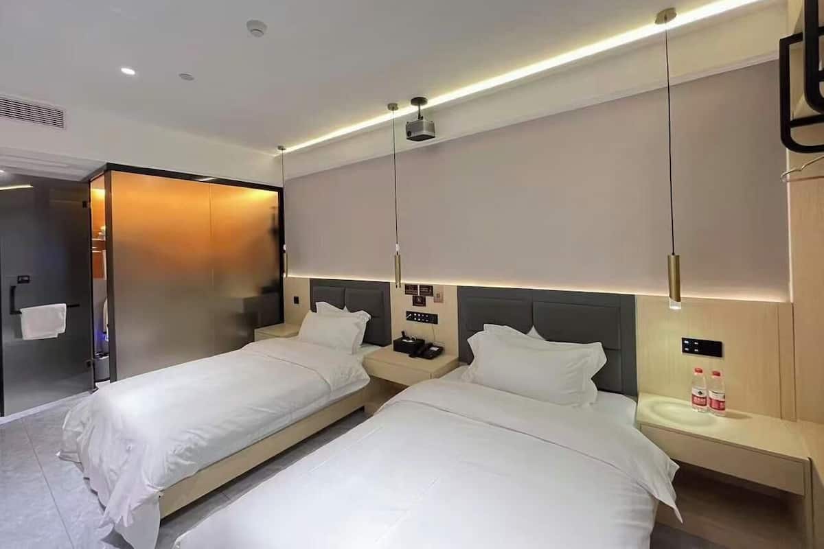 Quarto de hotel com duas camas, mesas de cabeceira, projetor fixado na parede e design moderno.