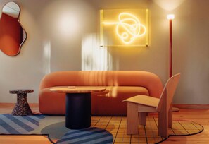 Quarto moderno com sofá, mesa redonda, cadeira, mesinha lateral e um letreiro de neon na parede.