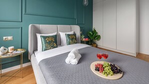 Cama bem arrumada com um edredom cinza, duas toalhas brancas dobradas e um prato de frutas com uvas e frutas vermelhas.