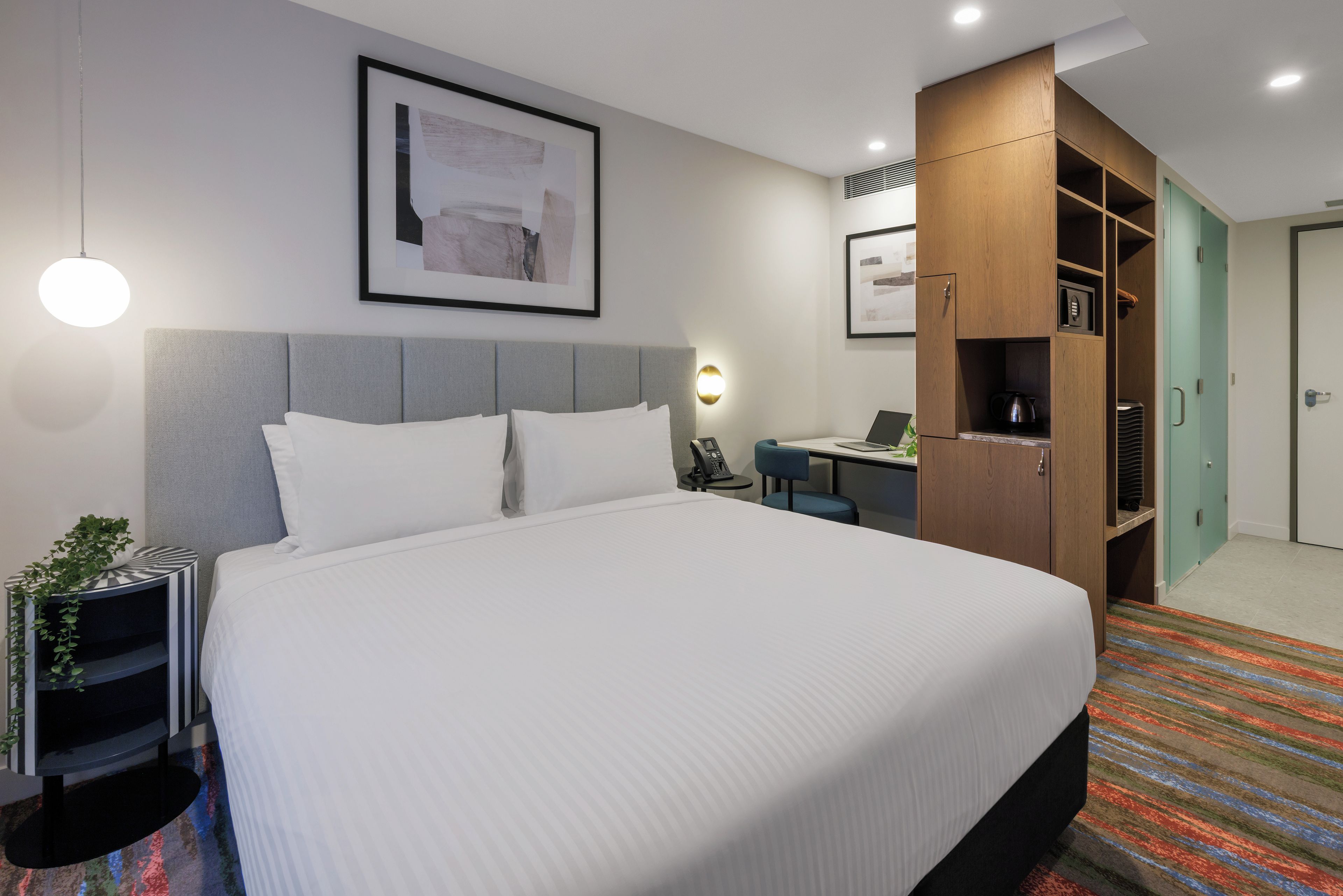 Quarto de hotel com cama, mesa de cabeceira, abajur, obra de arte emoldurada e armário embutido.