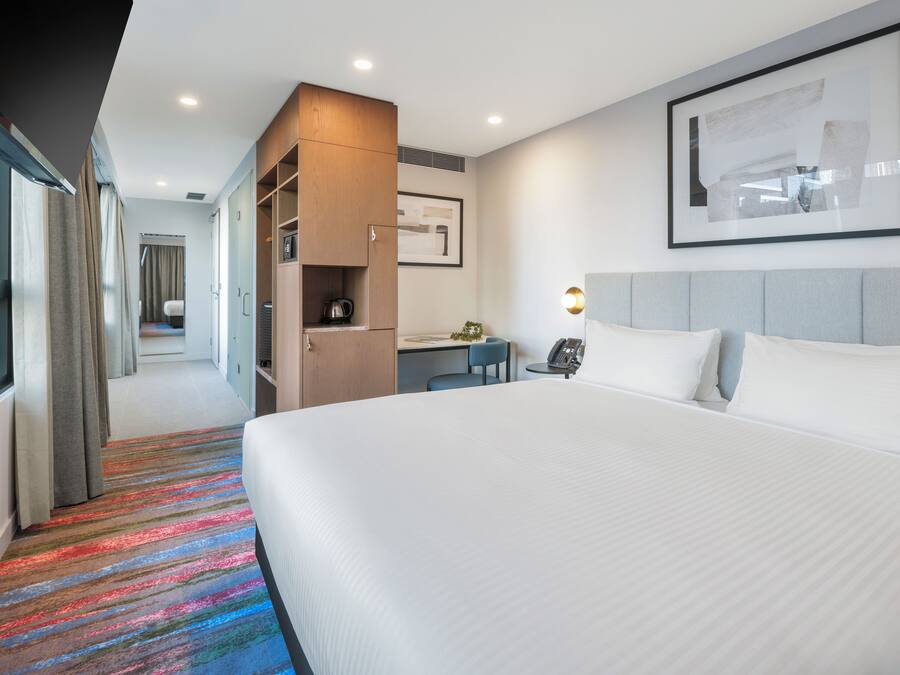 Quarto de hotel com uma cama grande, uma escrivaninha, um espaço com cozinha compacta e um banheiro visível através de uma porta aberta.