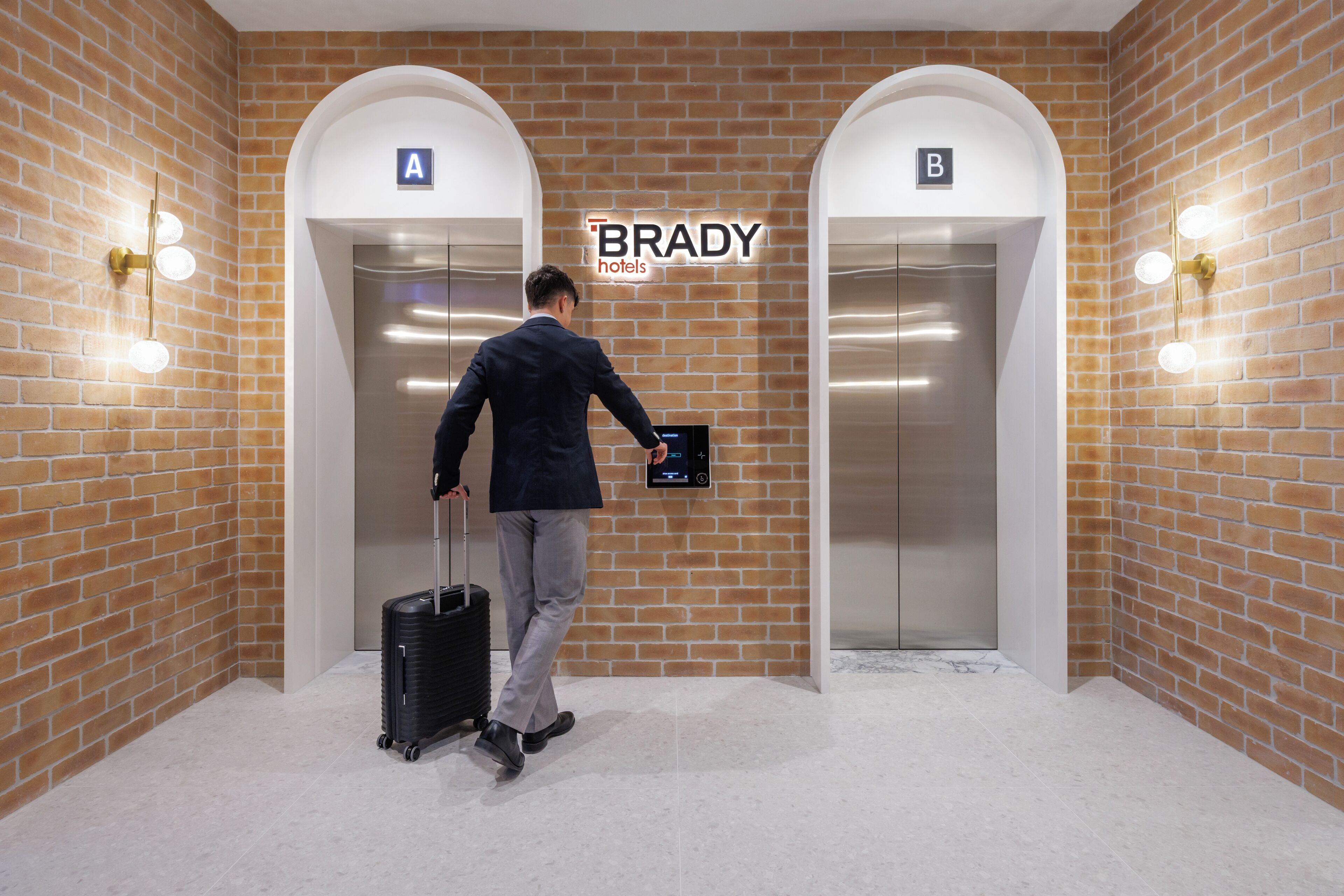 Um homem de terno com mala em um elevador com a sinalização 'BRADY hotels', paredes de tijolos e duas portas de elevador identificadas com as letras 'A' e 'B'.