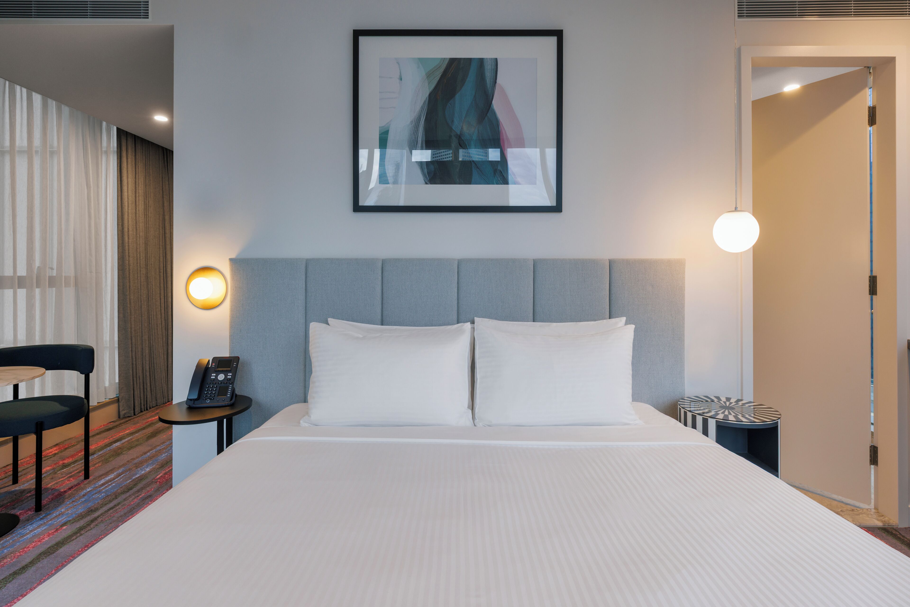 Quarto de hotel com uma cama, mesa de cabeceira com telefone, luminária na parede e uma obra de arte abstrata emoldurada.