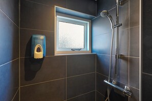 Casa de banho com azulejos escuros, janela, dispensador de sabonete líquido e chuveiro.