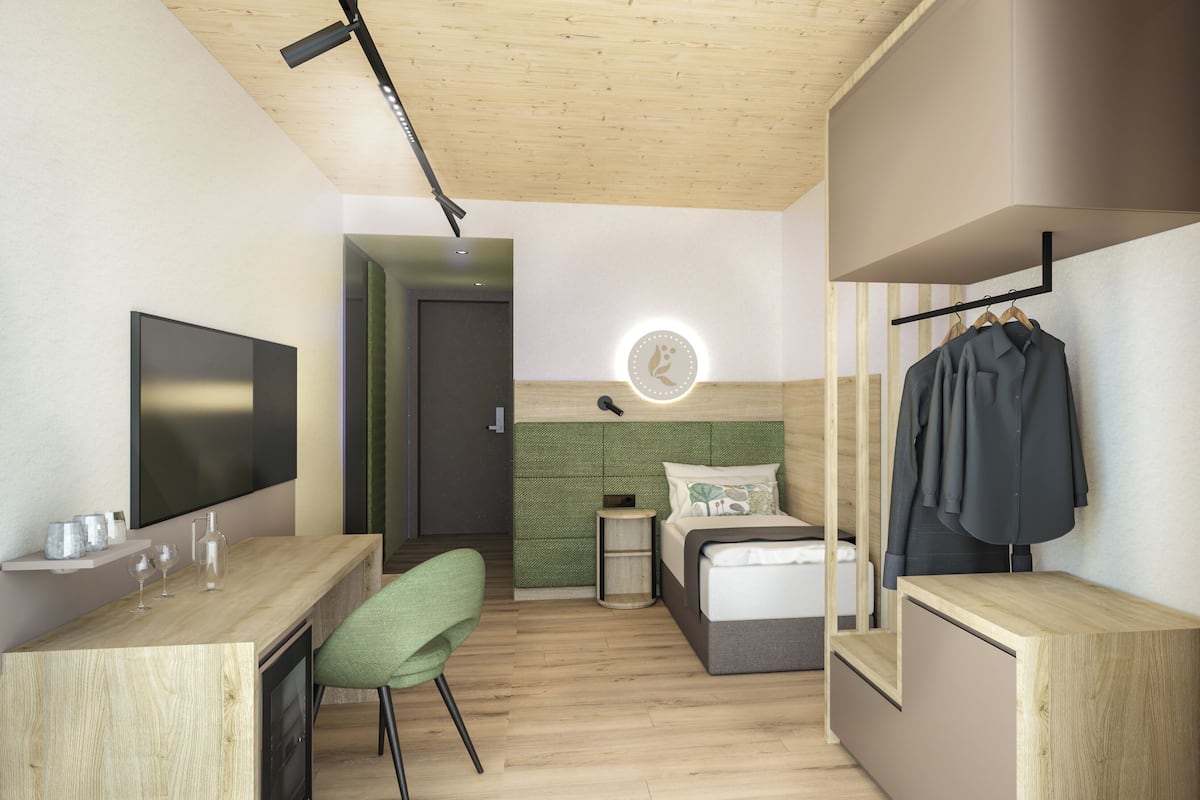 Quarto de hotel moderno com teto de madeira, TV de tela plana, escrivaninha com copos, cadeira verde, cama com cabeceira e um cabideiro.