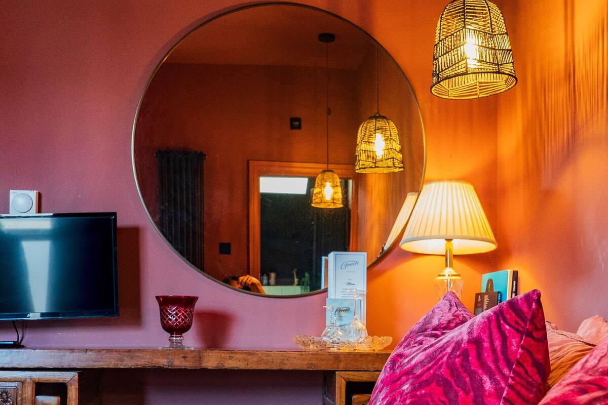 Quarto de hotel com uma cama, uma televisão, um espelho redondo, uma mesa de cabeceira de madeira e duas luminárias pendentes.