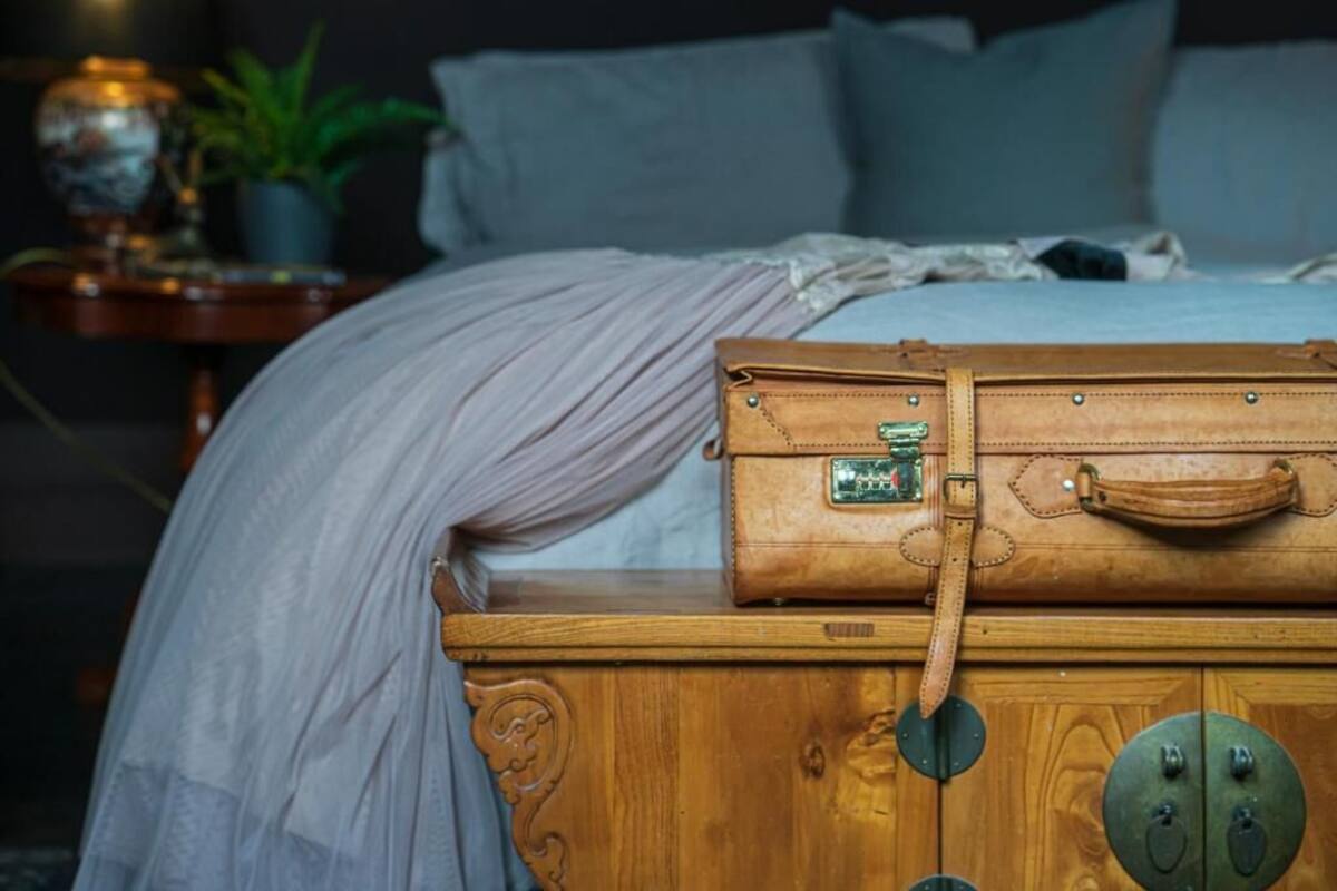 Uma cama com colcha branca, um baú de madeira com mala antiga, e uma mesinha com abajur e planta ao fundo.