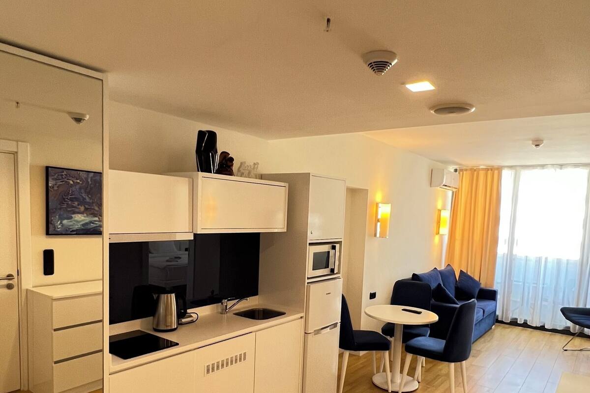 Cozinha moderna com armários brancos, micro-ondas e cafeteira. Possui um espaço de jantar com um sofá azul e uma pequena mesa. O quarto tem piso de madeira e uma grande janela com cortinas.