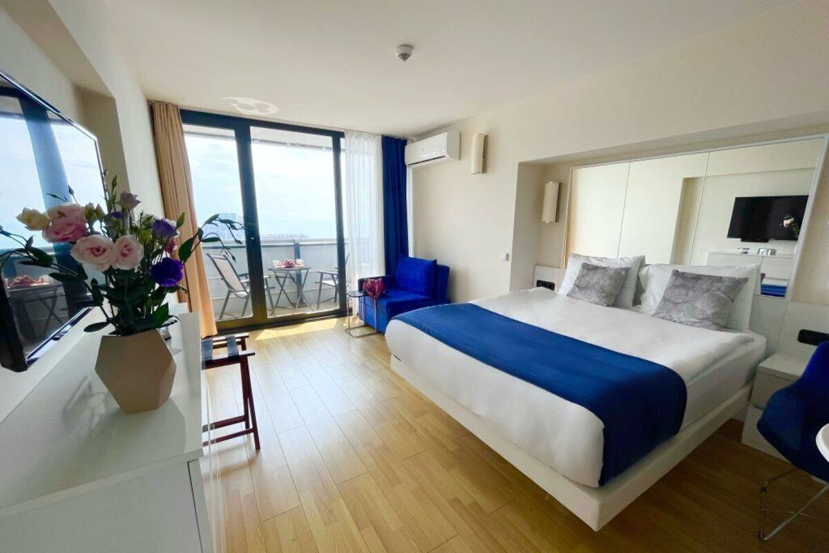 Quarto de hotel moderno com cama grande, varanda e vista, secretária com computador e televisão.