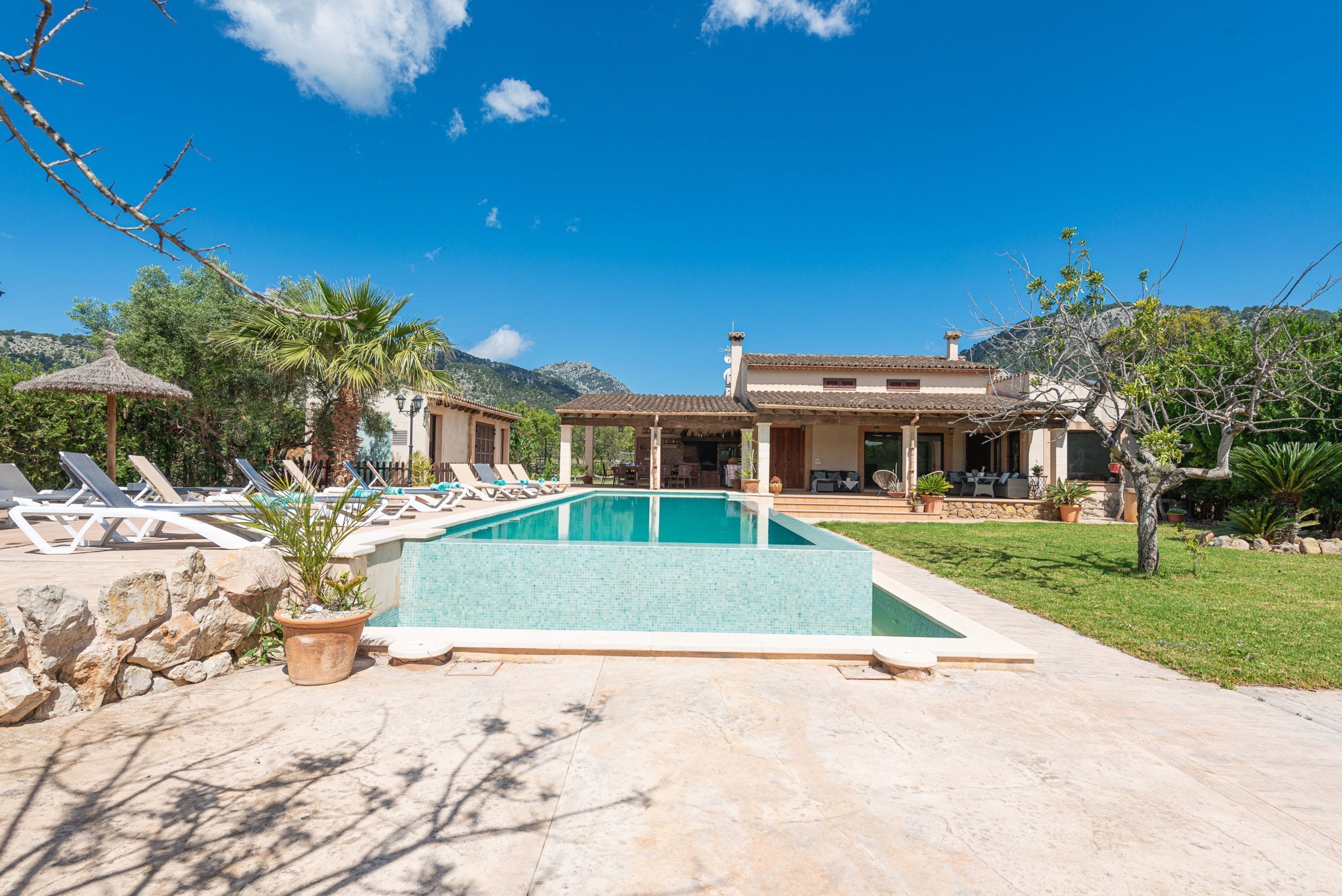 Uma piscina com espreguiçadeiras, uma casa com telhado em telhas e montanhas ao fundo.