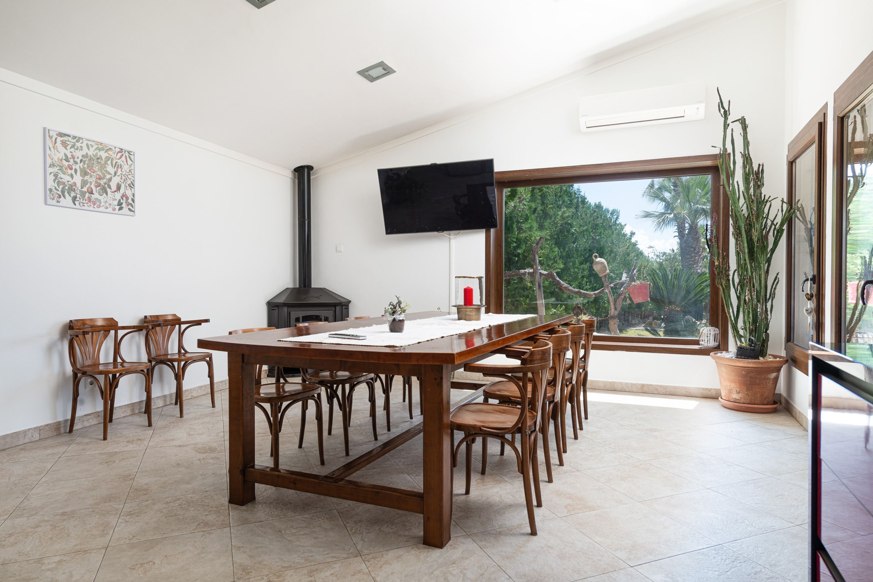Uma sala de jantar com uma mesa de madeira comprida, várias cadeiras, uma televisão na parede e uma grande janela com vista para o verde.