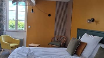 Quarto de hotel com uma cama, uma poltrona amarela, uma cadeira de vime, uma mesinha e uma janela com cortinas.