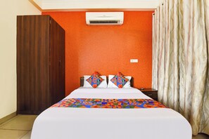 Quarto de hotel com parede laranja, uma cama com colcha geométrica colorida, duas almofadas com padrões geométricos, um guarda-roupa de madeira, uma mesa de cabeceira e uma janela com cortinas.