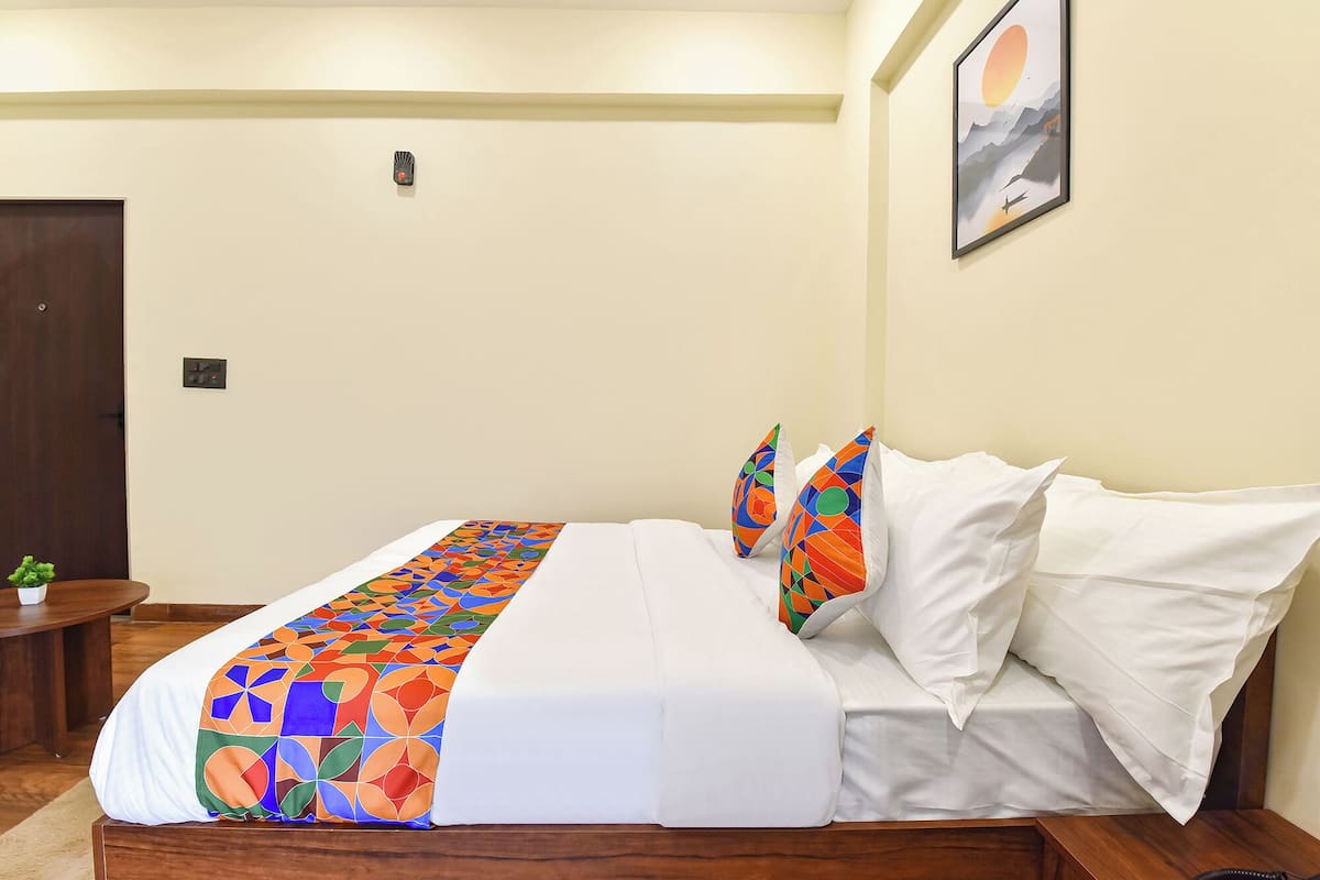 Cama bem arrumada com colcha de padrão geométrico colorido e almofadas brancas, cabeceira de madeira, mesa de cabeceira pequena com uma planta e um quadro na parede.