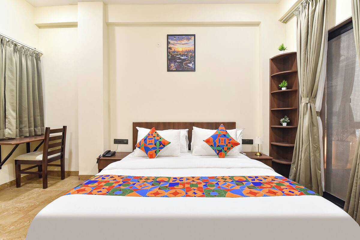 Quarto de hotel com cama, cadeira, mesa de cabeceira, luminária, quadro, prateleira com plantas e janela com cortinas.