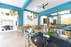 Uma área de refeições com móveis de vime, uma mesa central com um vaso, ventiladores de teto e uma parede com quadros e plantas.
