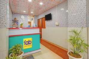 Uma área de recepção de hotel com parede estampada, balcão de atendimento, relógio, televisão e plantas em vasos.