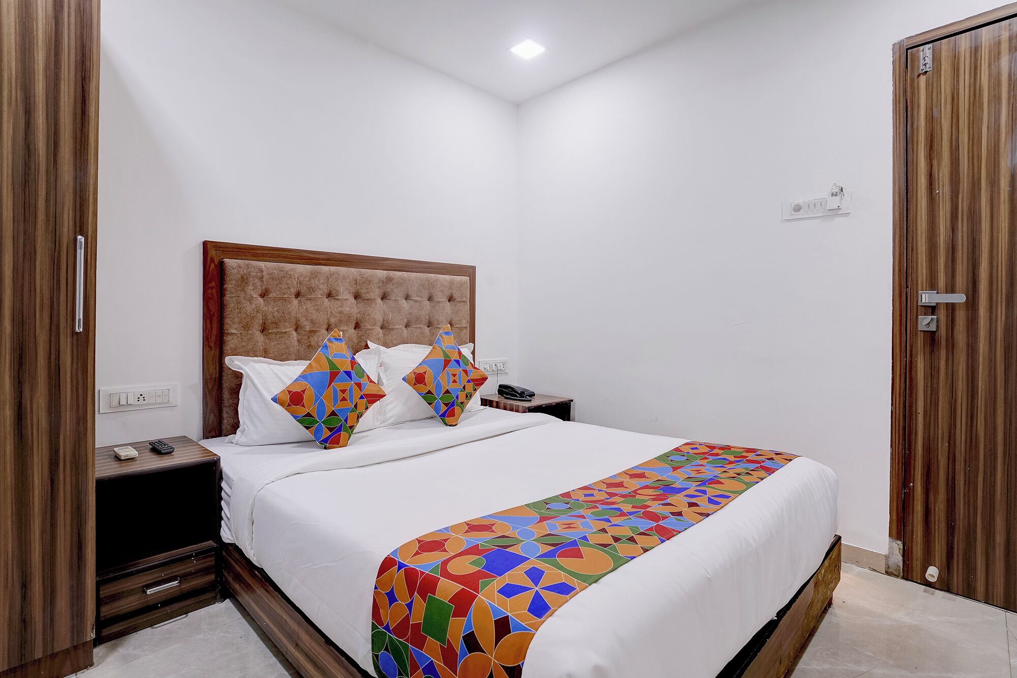 Quarto com cabeceira estofada, cama com roupa de cama branca, edredom colorido com padrão geométrico, mesa de cabeceira com abajur e guarda-roupa de madeira.