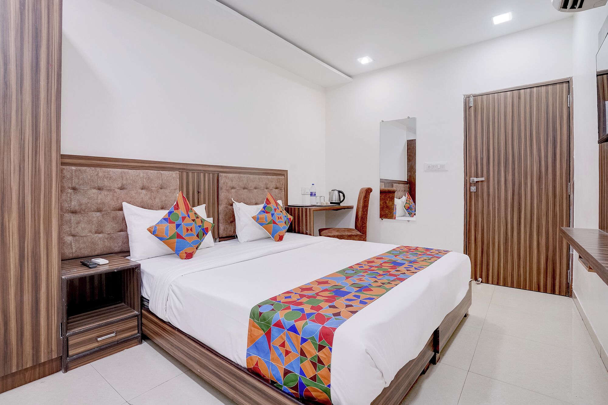 Quarto de hotel com cama, mesa de cabeceira, escrivaninha, cadeira, espelho e guarda-roupa de madeira.