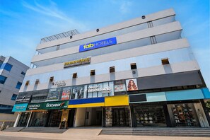 Um hotel de vários andares com design moderno, exibindo uma placa chamativa para 'Shabari Heights Luxury Abode NC Rooms' e diversas lojas no andar térreo.