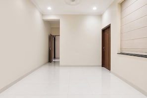 Um corredor com piso de azulejos brancos, uma porta de madeira à direita e uma luminária embutida no teto.