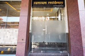 Entrada de um edifício com uma placa indicando 'Zamzam Residency' e uma porta de vidro com persianas horizontais.