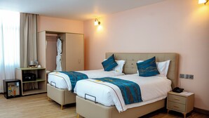 Quarto de hotel com duas camas, um guarda-roupa, uma mesa de cabeceira com telefone e um pequeno armário.