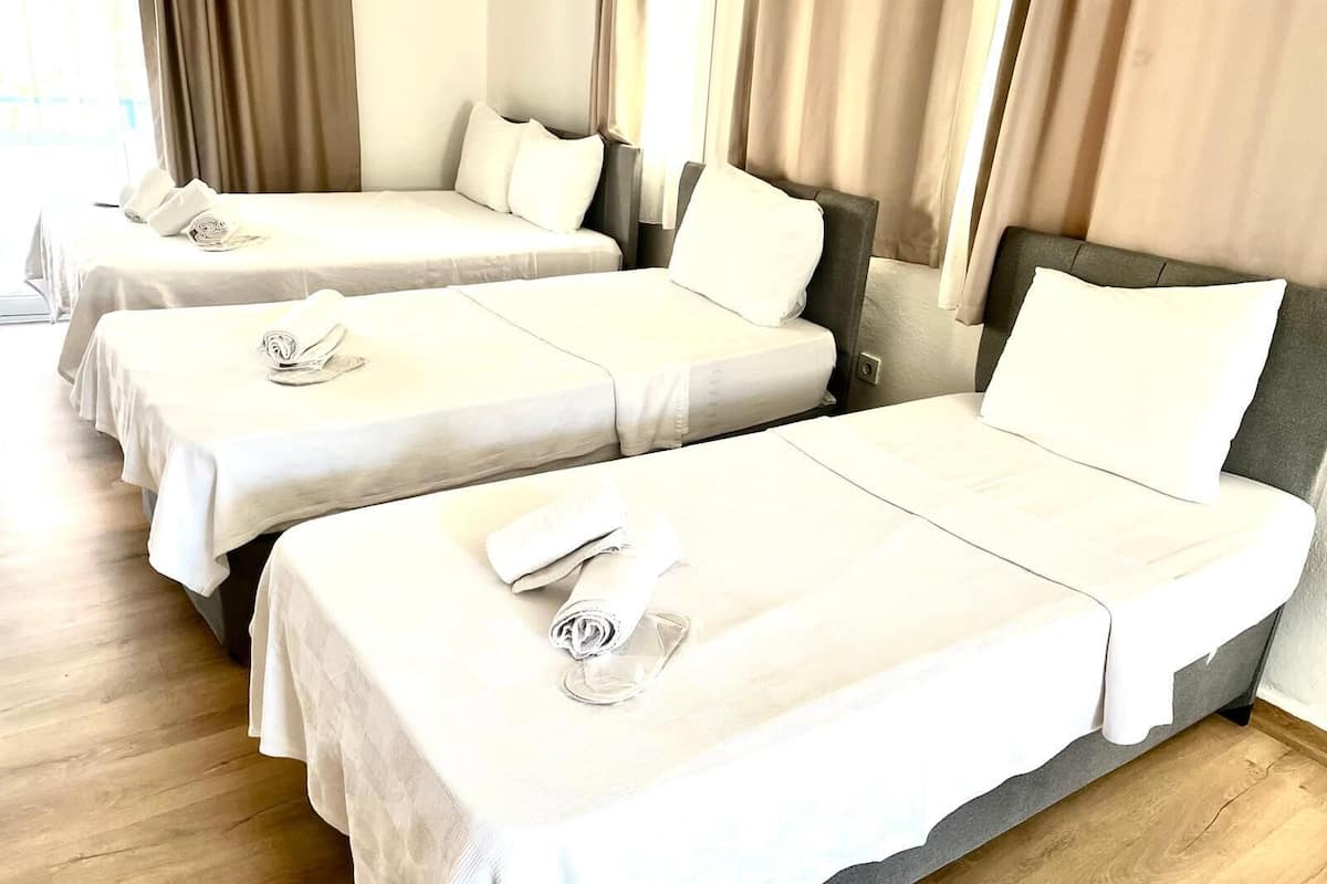 Quarto de hotel com três camas de solteiro, cada uma com colcha e travesseiros brancos, e piso de madeira.
