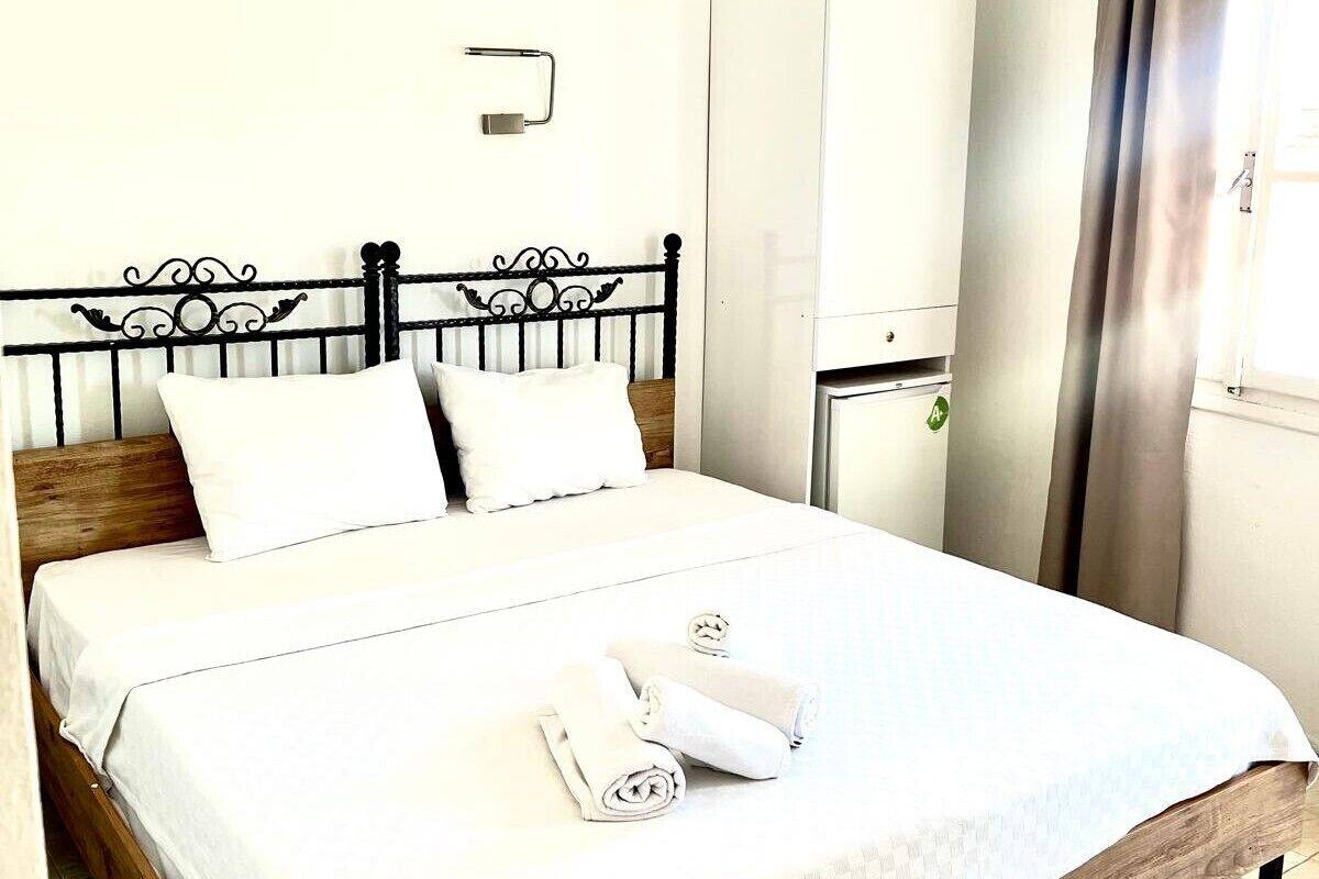 Cama bem arrumada com lençóis brancos e toalhas dobradas, cabeceira de madeira e parede branca com porta e interruptor de luz.