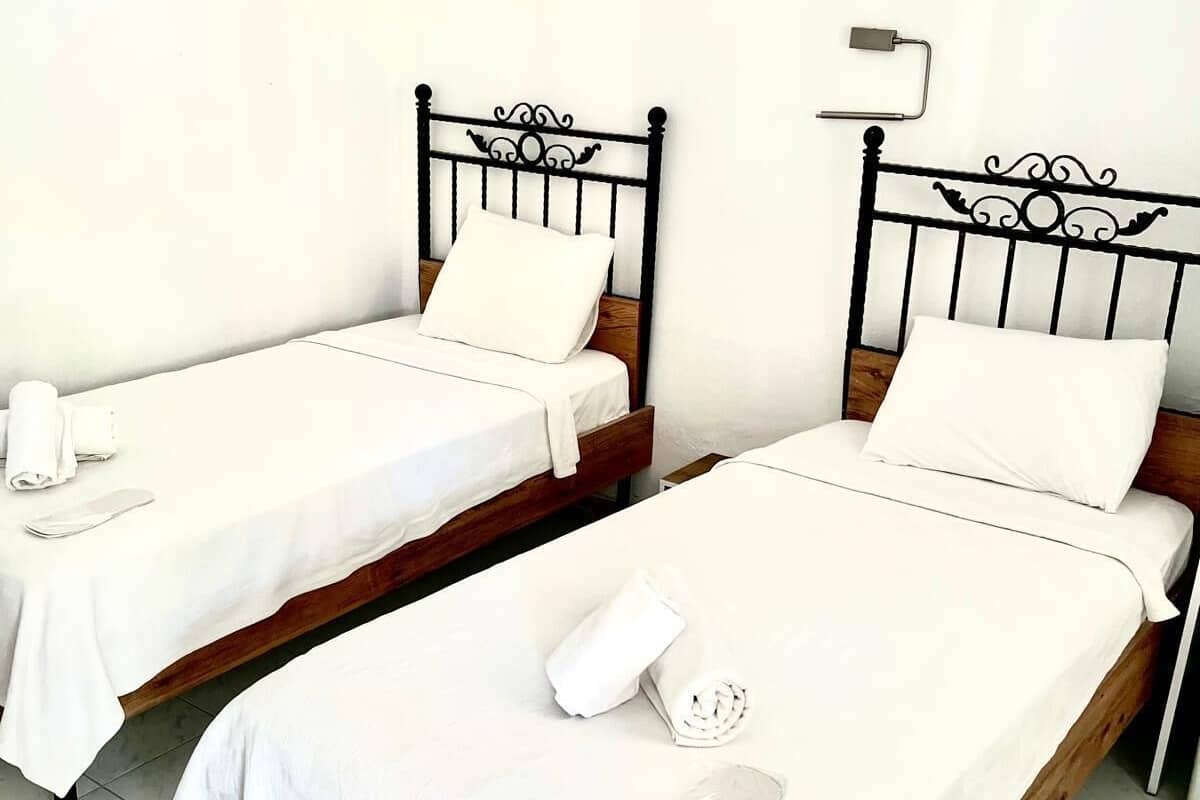 Duas camas de solteiro com estrutura de madeira e roupa de cama branca, um ar condicionado fixado na parede e uma parede branca lisa.
