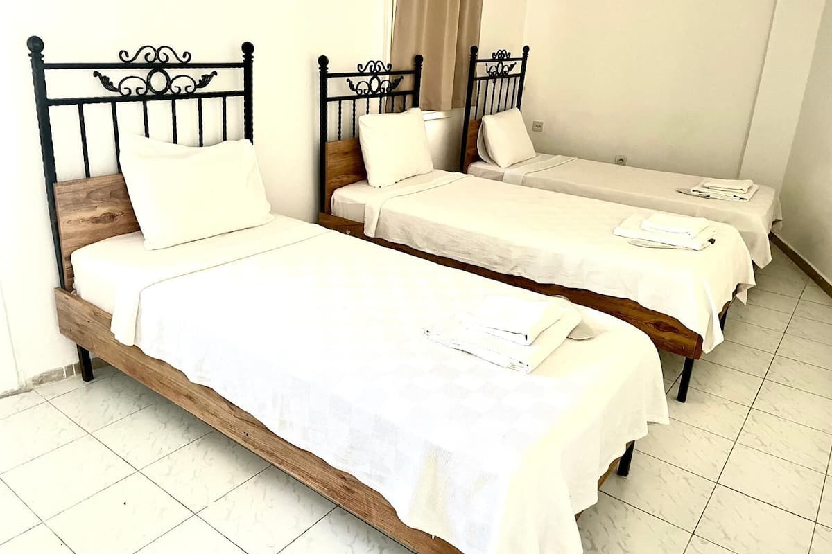 Três camas de solteiro com estruturas de madeira e roupa de cama branca em um quarto com piso ladrilhado.