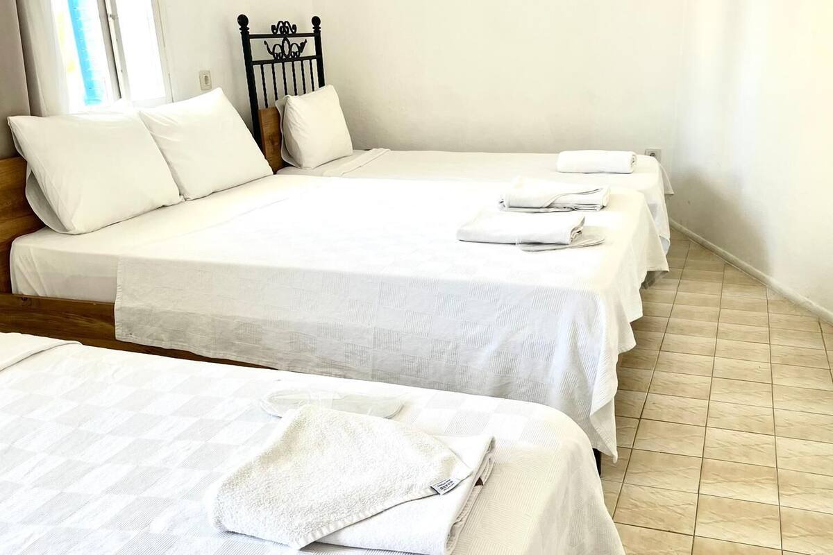 Quarto de hotel com três camas, cada uma com roupa de cama e almofadas brancas, cabeceira de madeira e piso ladrilhado.