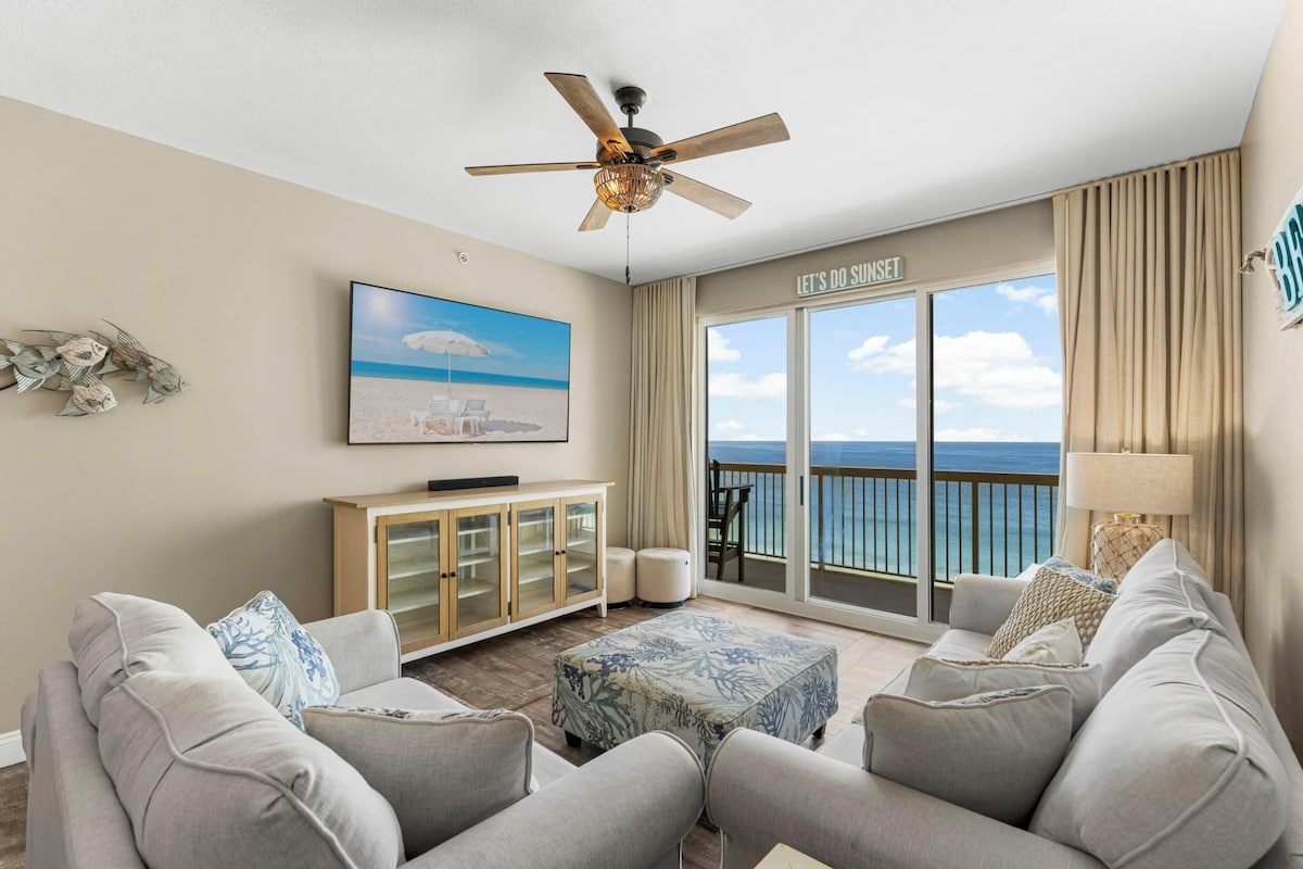 Uma sala de estar com televisão, ventilador de teto, sofá, mesa de centro e vista para o mar através de grandes janelas.