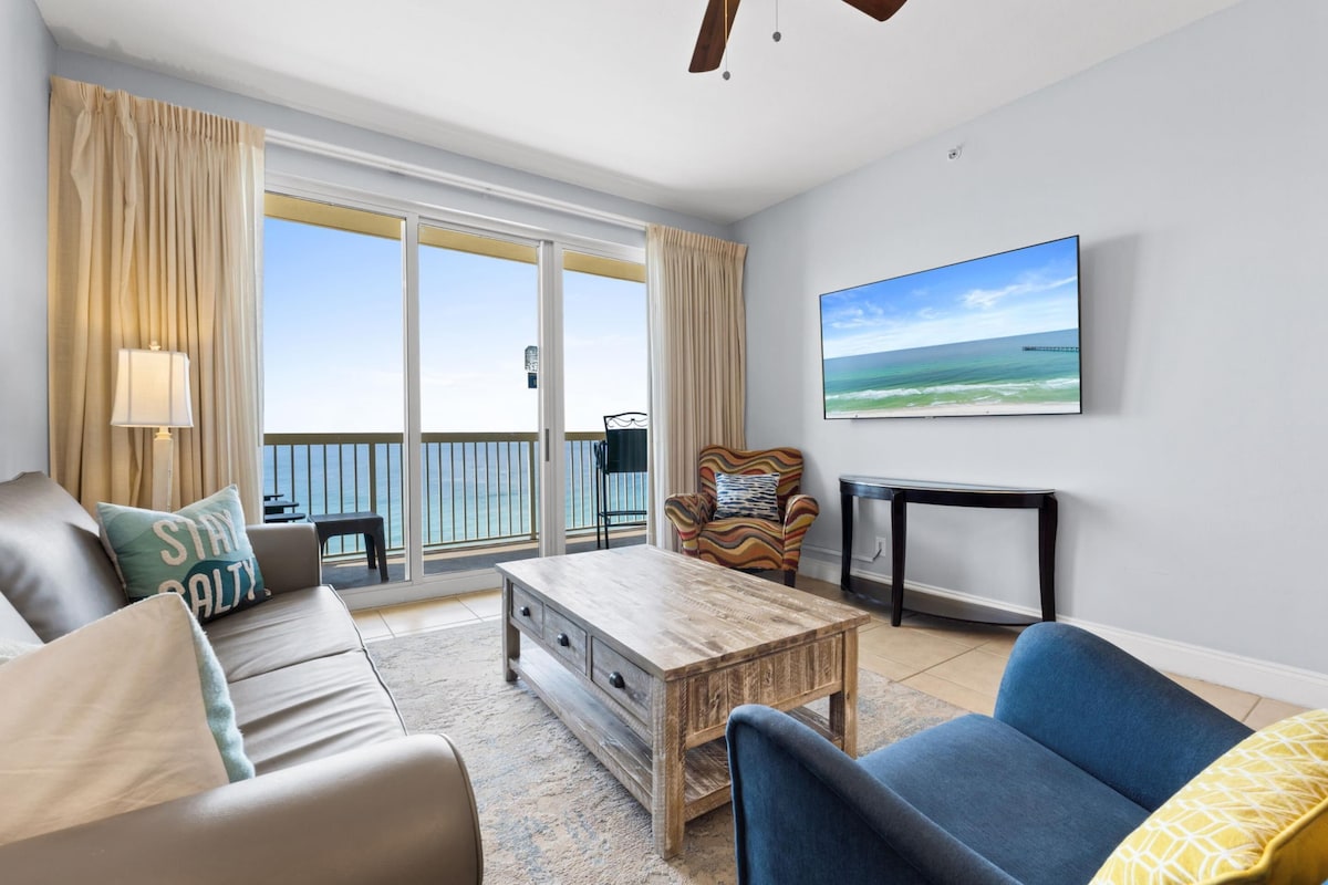 Uma sala de estar espaçosa com sofá, poltrona, mesa de centro e TV na parede. Varanda com vista para o oceano visível através de grandes portas deslizantes.