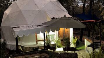 Uma tenda em forma de abóbada com exterior branco e interior cinzento, rodeada por um jardim com diversas plantas e um pequeno lago.