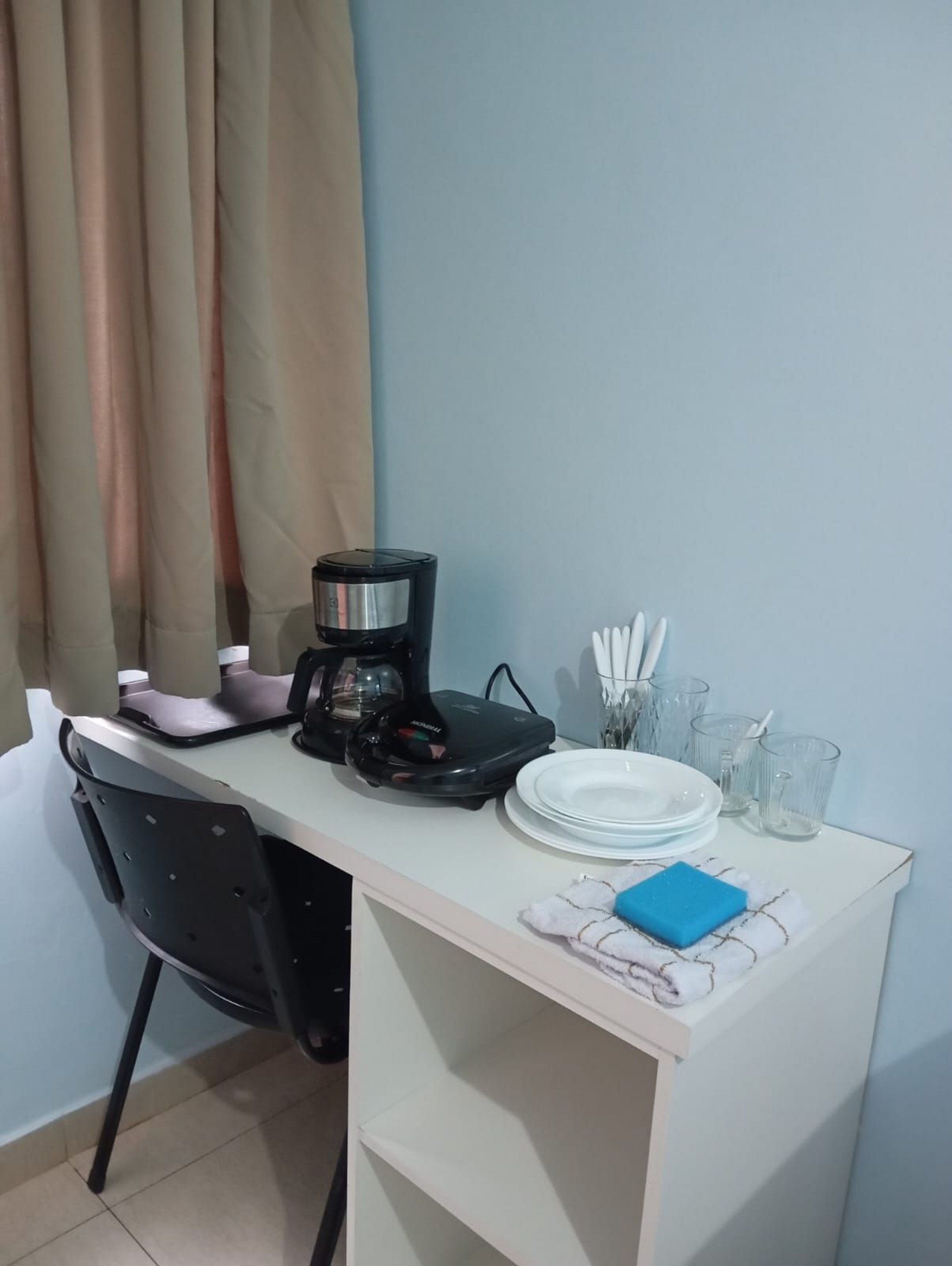 Uma pequena mesa branca com uma máquina de café preta, uma pilha de pratos brancos, uma esponja azul e um pano branco.