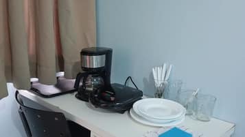 Uma pequena mesa branca com uma máquina de café preta, uma pilha de pratos brancos, uma esponja azul e um pano branco.